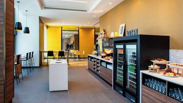 Um café moderno com um balcão central branco, área de estar com cadeiras, cozinha com refrigerador cheio de bebidas e um buffet com frutas e doces.