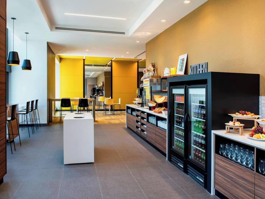 Um café moderno com um balcão central branco, área de estar com cadeiras, cozinha com refrigerador cheio de bebidas e um buffet com frutas e doces.