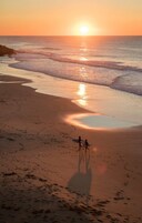 Cena de praia com pôr do sol, ondas e duas pessoas caminhando com pranchas de surf.