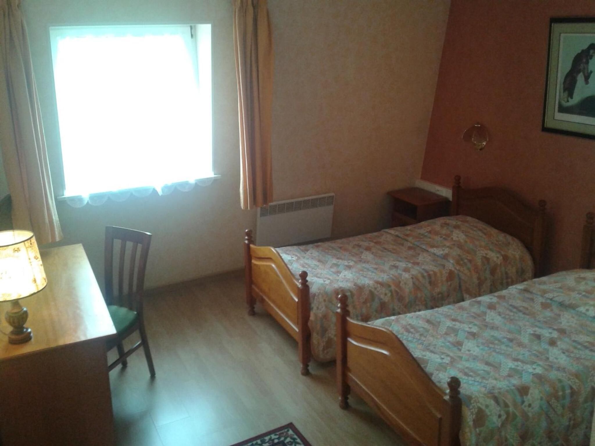 Um quarto pequeno e modesto, com duas camas de madeira, uma escrivaninha, uma cadeira e uma janela com cortinas.