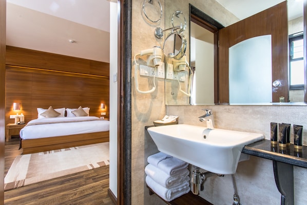 Quarto de hotel com cama, banheiro com pia e espelho, e mesa de cabeceira com abajur.