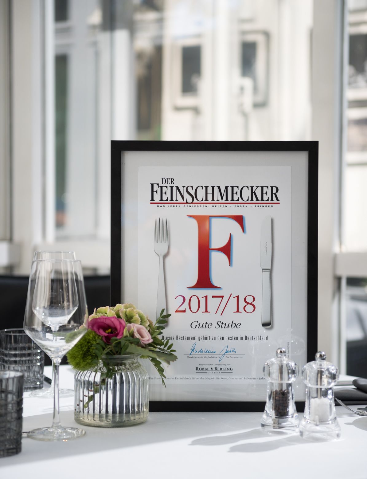 Cardápio emoldurado intitulado "Der Feinschmecker 2017/18" com uma grande letra "F" vermelha e ilustração de garfo e faca.