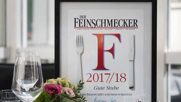Cardápio emoldurado intitulado "Der Feinschmecker 2017/18" com uma grande letra "F" vermelha e ilustração de garfo e faca.