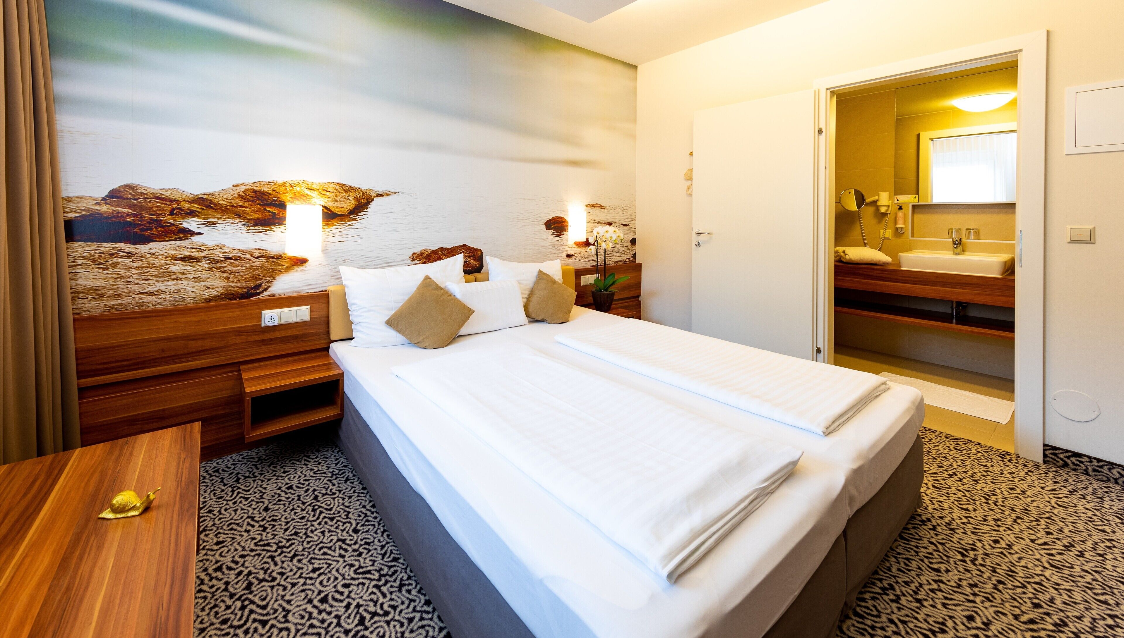 Quarto de hotel com cama, mesa de cabeceira, abajur, espelho e banheiro visível através de uma porta aberta.