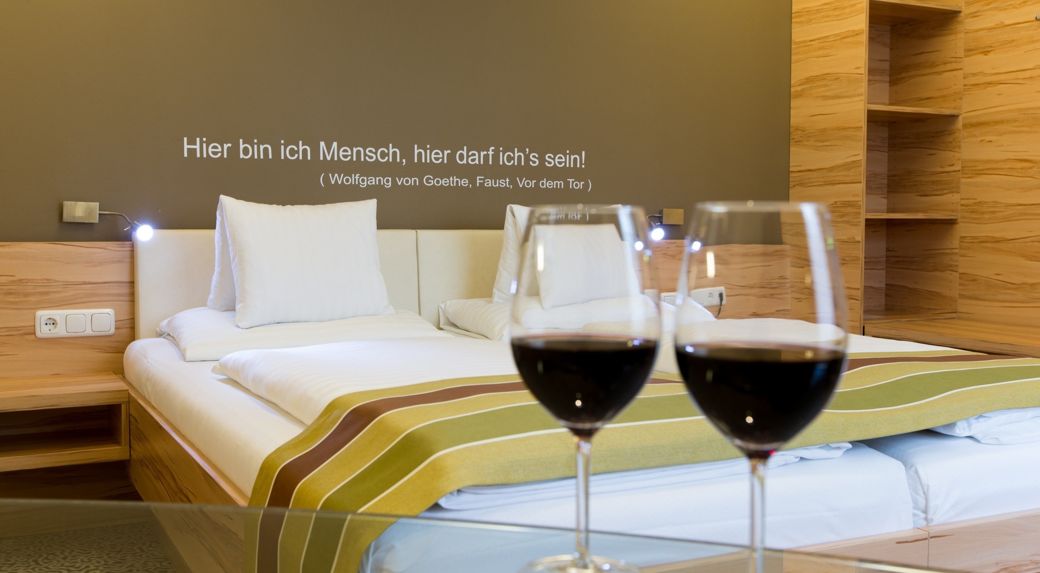 Quarto de hotel com uma cama, dois copos de vinho e uma frase na parede.