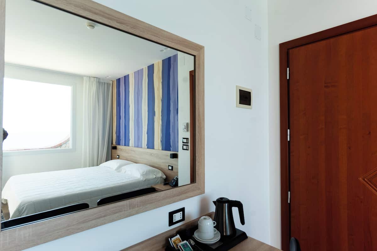 Quarto de hotel com uma cama, um espelho grande, uma porta de madeira e uma cafeteira no criado-mudo.
