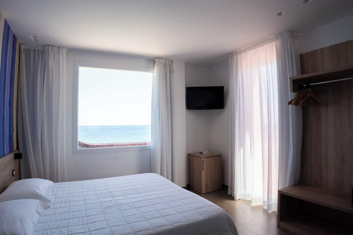Quarto de hotel com cama, TV fixada na parede, armário de madeira e janela com vista para o mar.