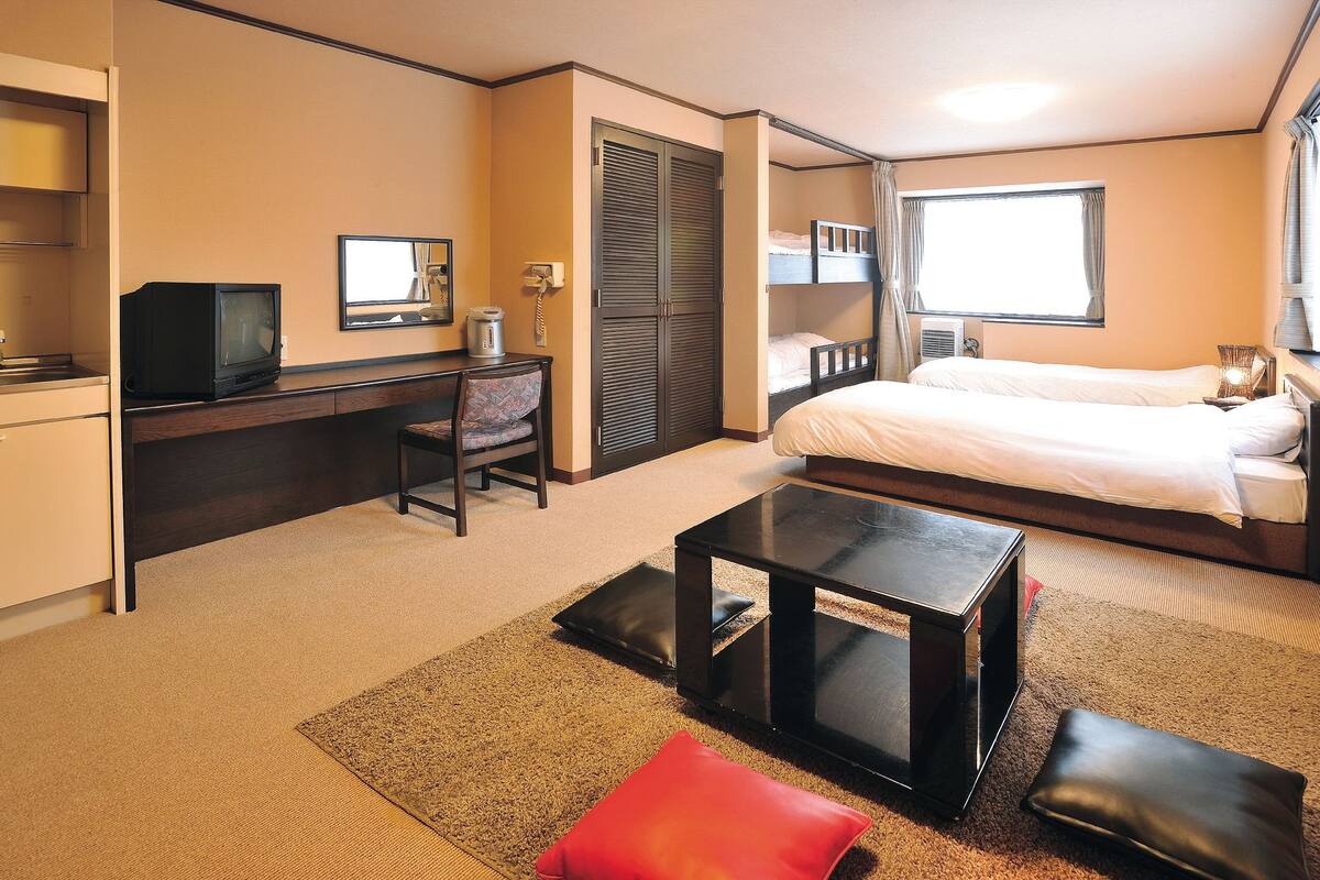 Quarto de hotel com cama, escrivaninha com televisão, cadeira, mesa pequena e janela.