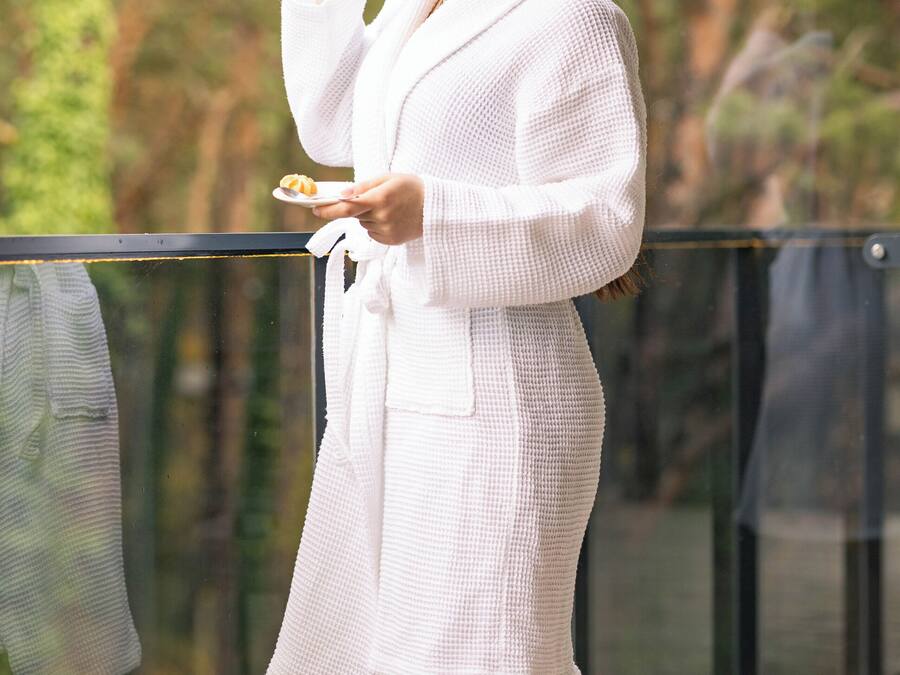 Uma pessoa vestindo um robe branco está em uma varanda com corrimão de vidro, segurando uma xícara e um prato.