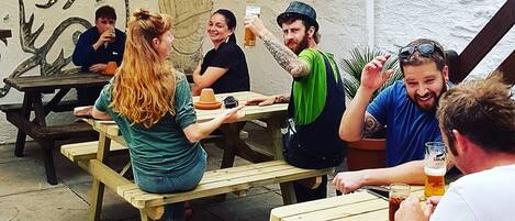 Um grupo de pessoas sentadas em mesas de madeira ao ar livre, conversando e bebendo cerveja.