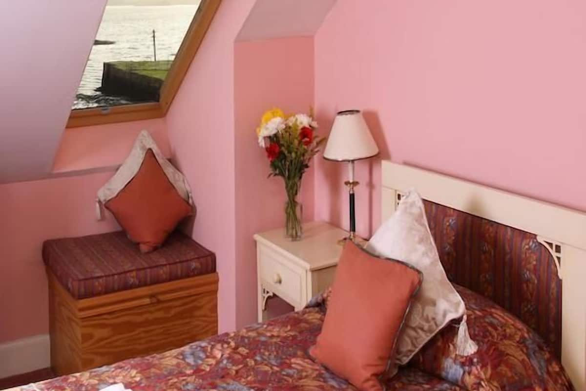 Um quarto com claraboia, uma cama com roupa de cama floral, uma mesa de cabeceira, um abajur e um vaso com flores.