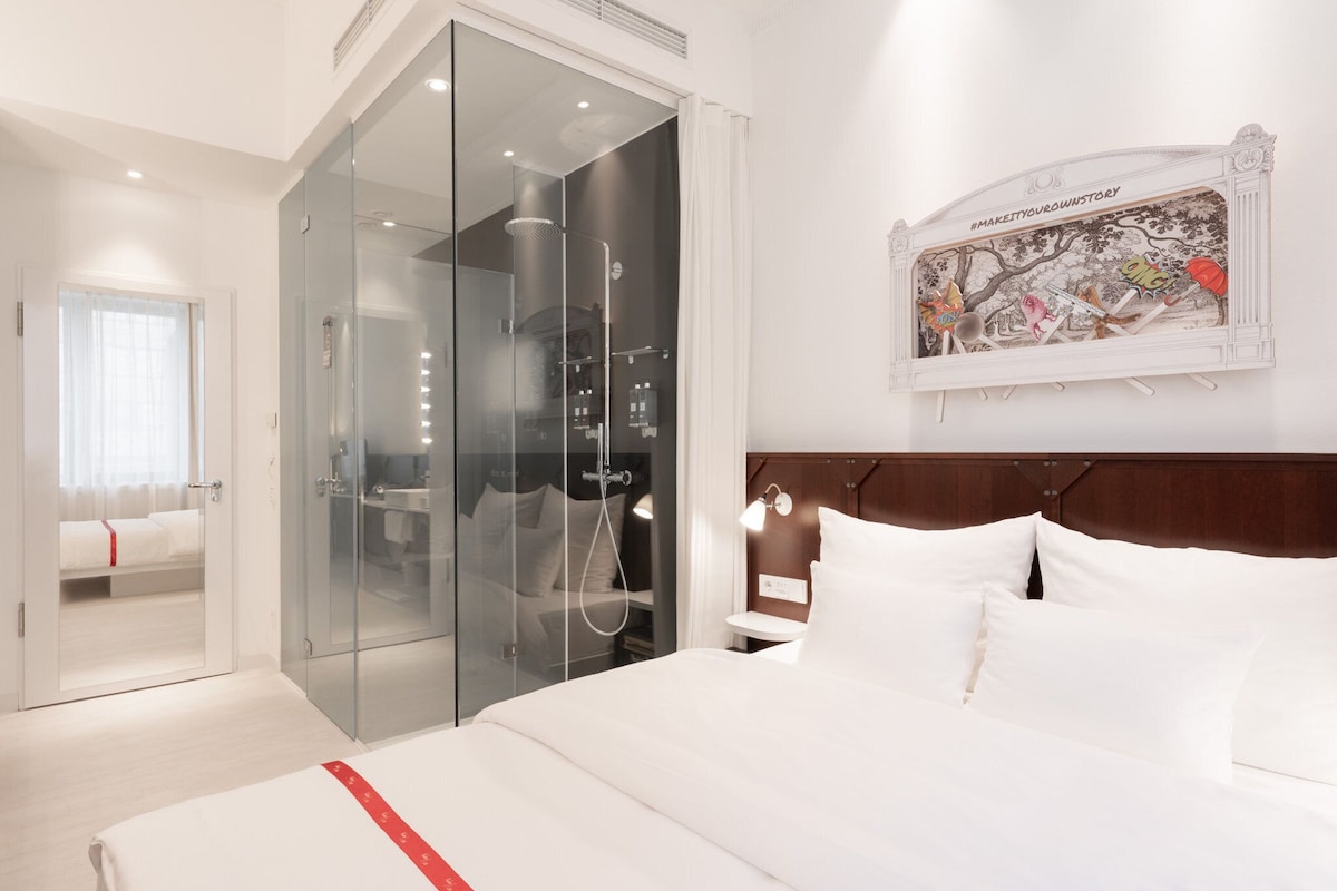 Quarto de hotel moderno com uma cama grande, closet envidraçado e espaço para chuveiro, além de uma obra de arte fixada na parede.