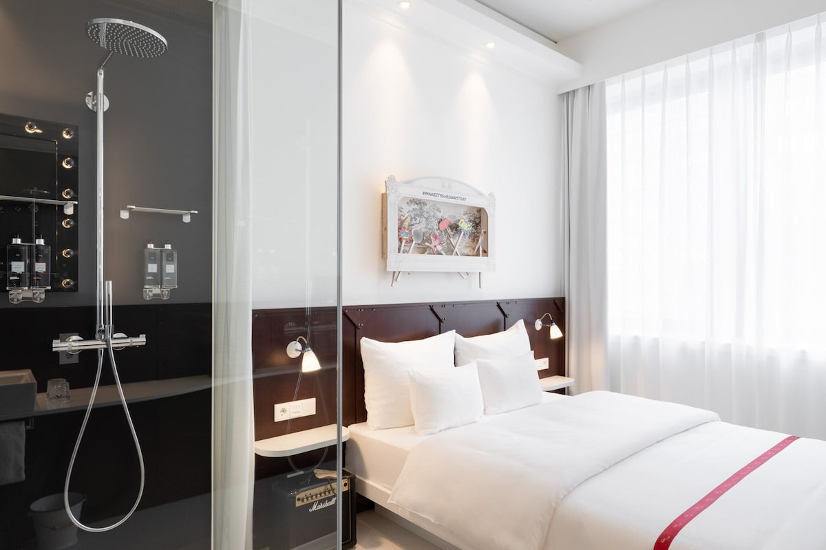 Quarto de hotel com cama, chuveiro, penteadeira com espelho e um quadro na parede.