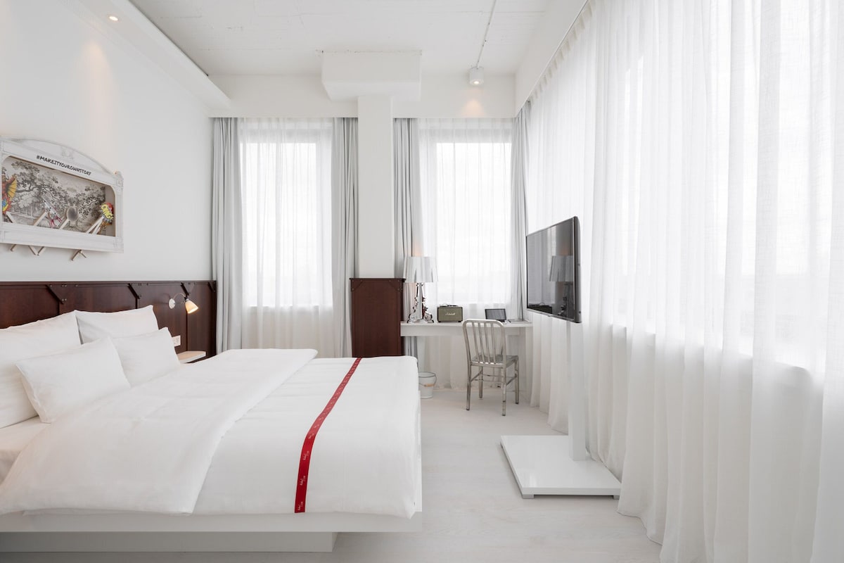 Quarto de hotel com uma cama grande, uma escrivaninha, uma cadeira, uma televisão e um quadro na parede.