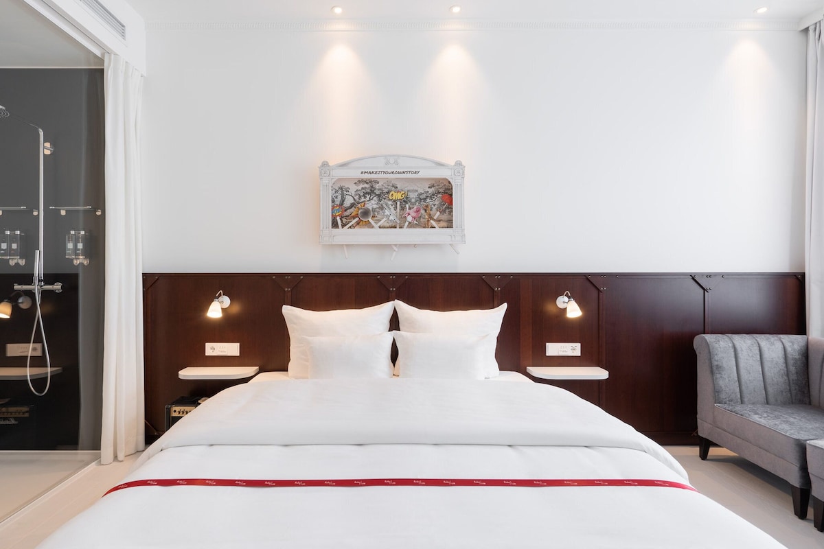 Quarto de hotel com uma cama grande, cabeceira com luzes fixadas na parede, mesa de cabeceira e uma poltrona cinza.