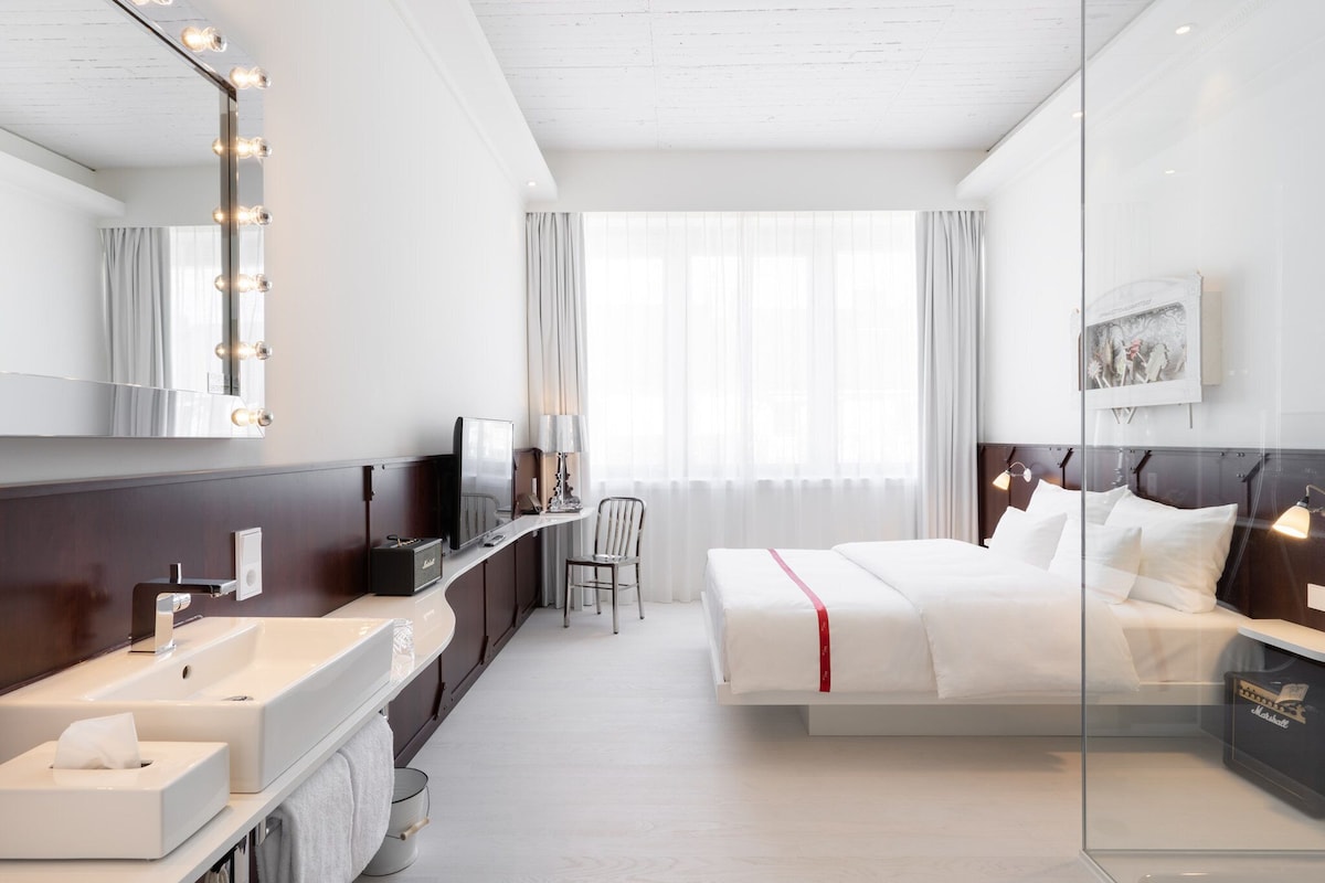 Quarto de hotel moderno com uma cama grande, uma toucador com espelho e iluminação, uma televisão e uma janela ampla com cortinas.