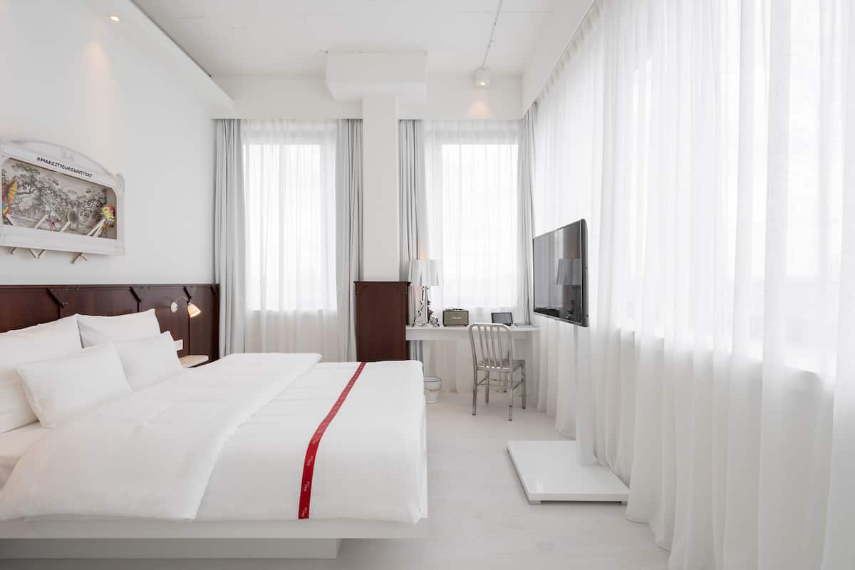 Quarto de hotel com cama, escrivaninha, cadeira, televisão e cortinas.