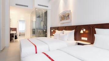 Quarto de hotel com duas camas, cabeceira de madeira, mesa de cabeceira pequena, luminária fixada na parede e box de vidro.