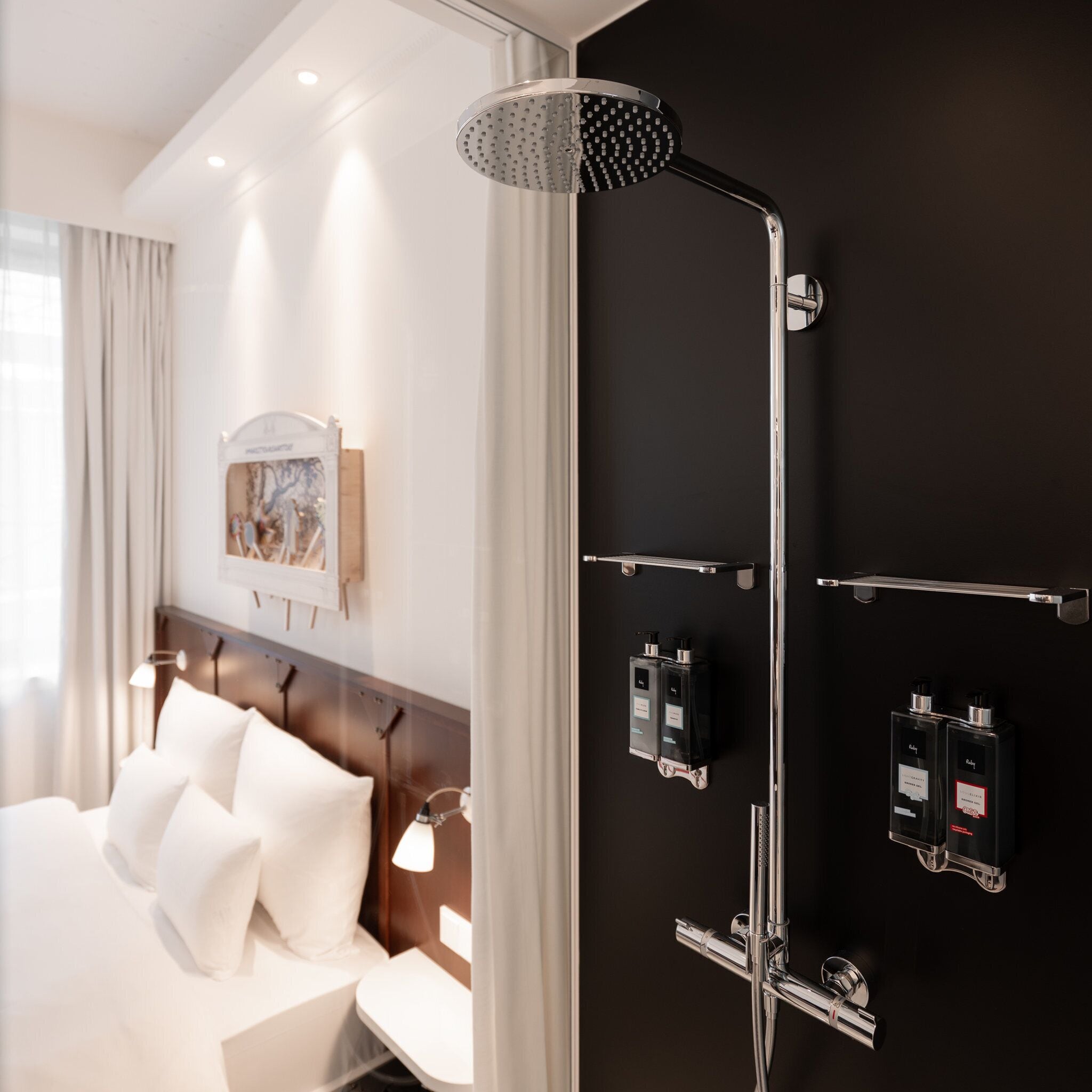 Quarto de hotel moderno com uma cama grande, chuveiro com cabeça de chuveiro tipo chuva e duas embalagens de produtos de higiene pessoal na parede.