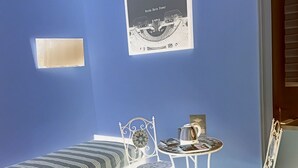 Quarto com parede azul, cama com roupa de cama listrada, mesinha com chaleira e uma cadeira.
