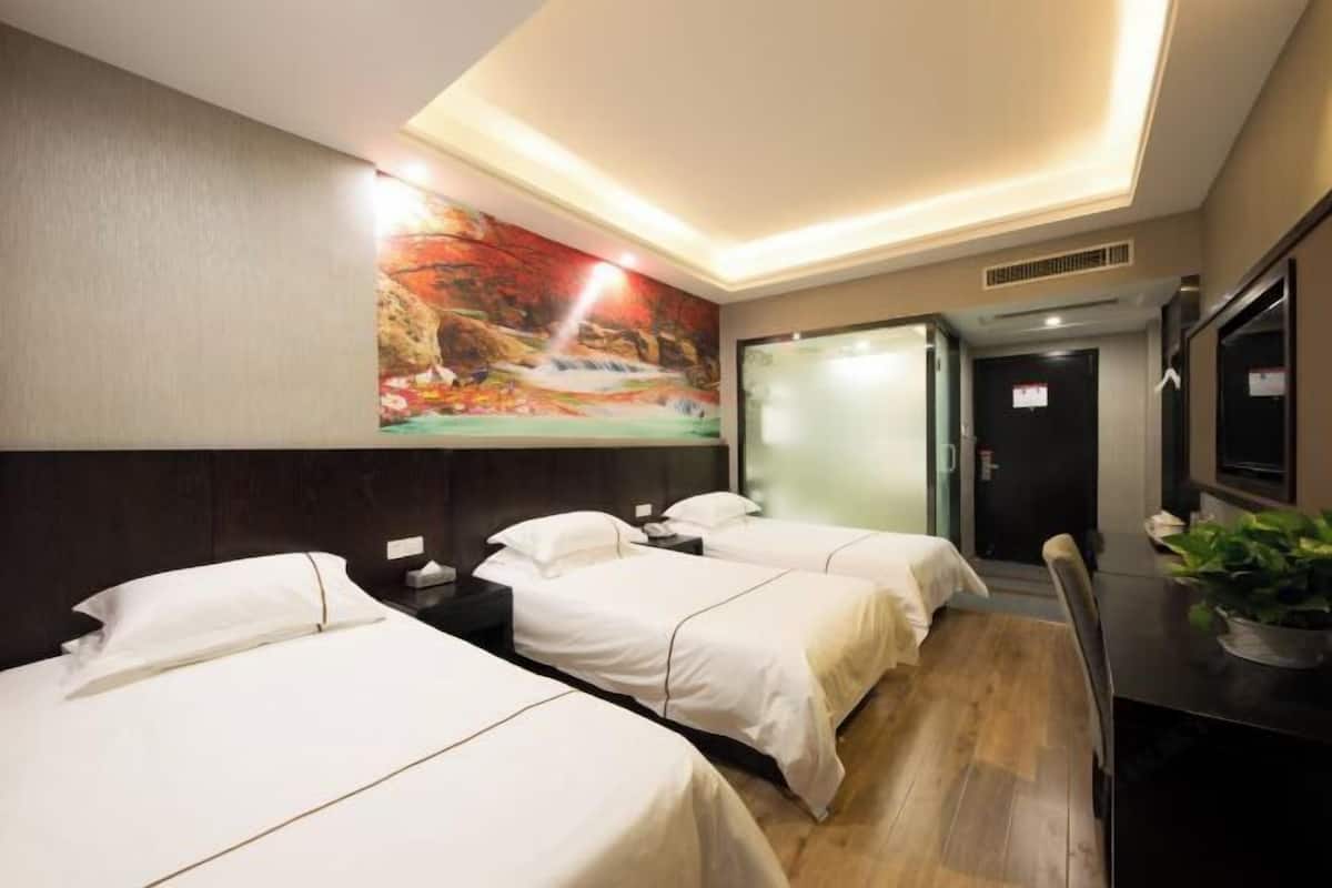 Quarto de hotel com três camas, piso de madeira, um quadro na parede e porta para o banheiro.