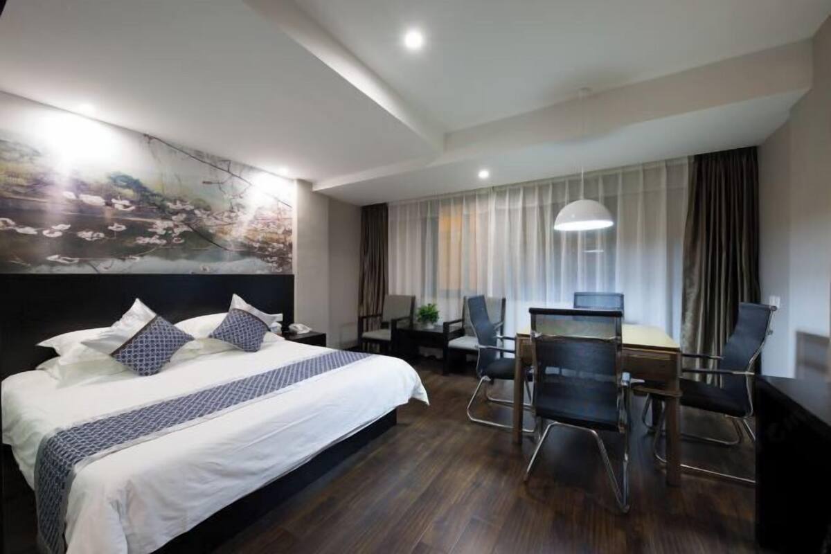 Quarto de hotel com uma cama grande, uma escrivaninha com cadeiras, um quadro na parede e uma janela com cortinas.