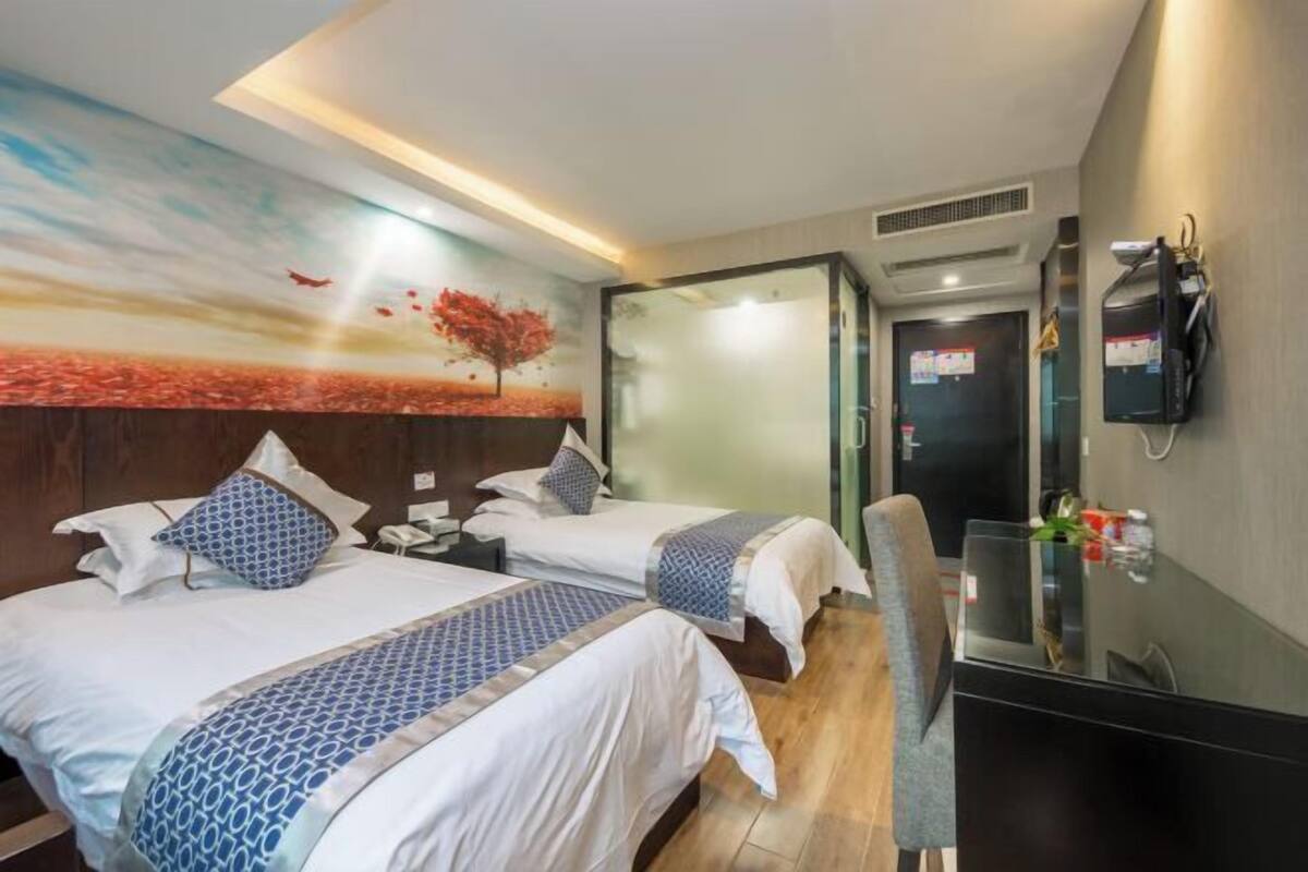Quarto de hotel com duas camas, uma escrivaninha, uma cadeira, uma televisão e um quadro na parede.
