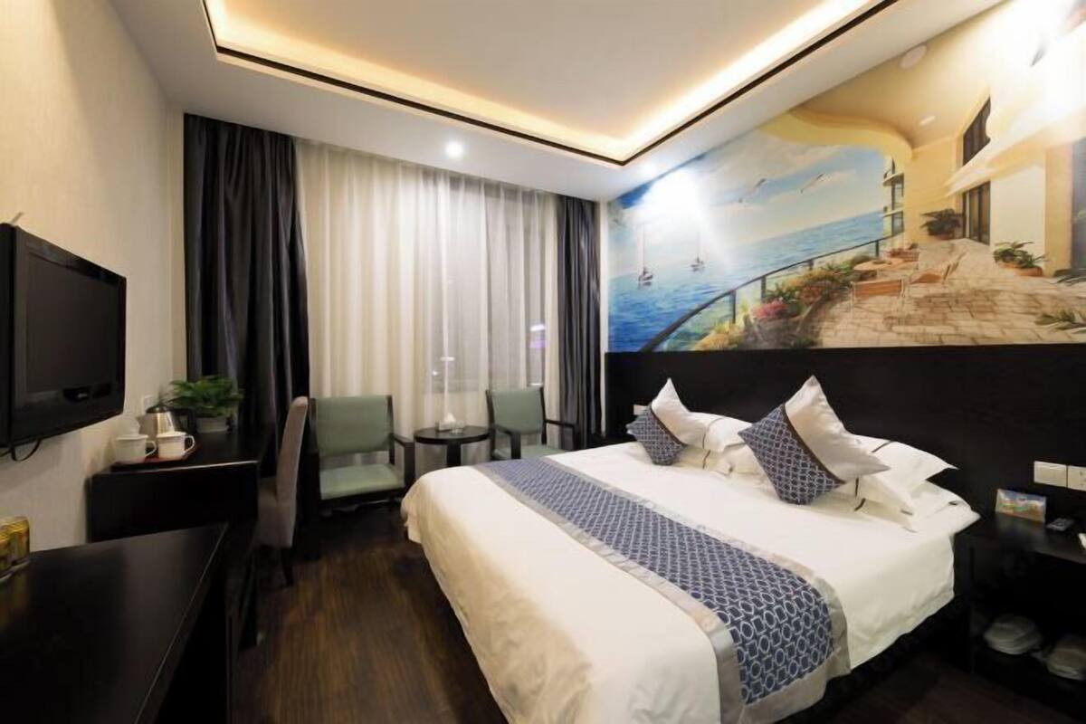 Quarto de hotel com cama, secretária, cadeira, televisão e pintura de uma paisagem costeira.