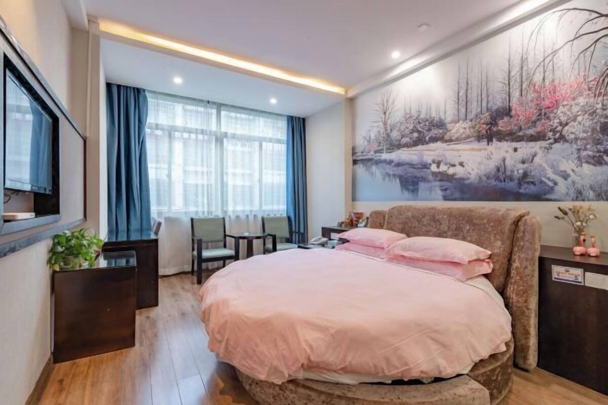 Quarto com cama, televisão, escrivaninha, cadeira, janela com cortinas e um quadro na parede.