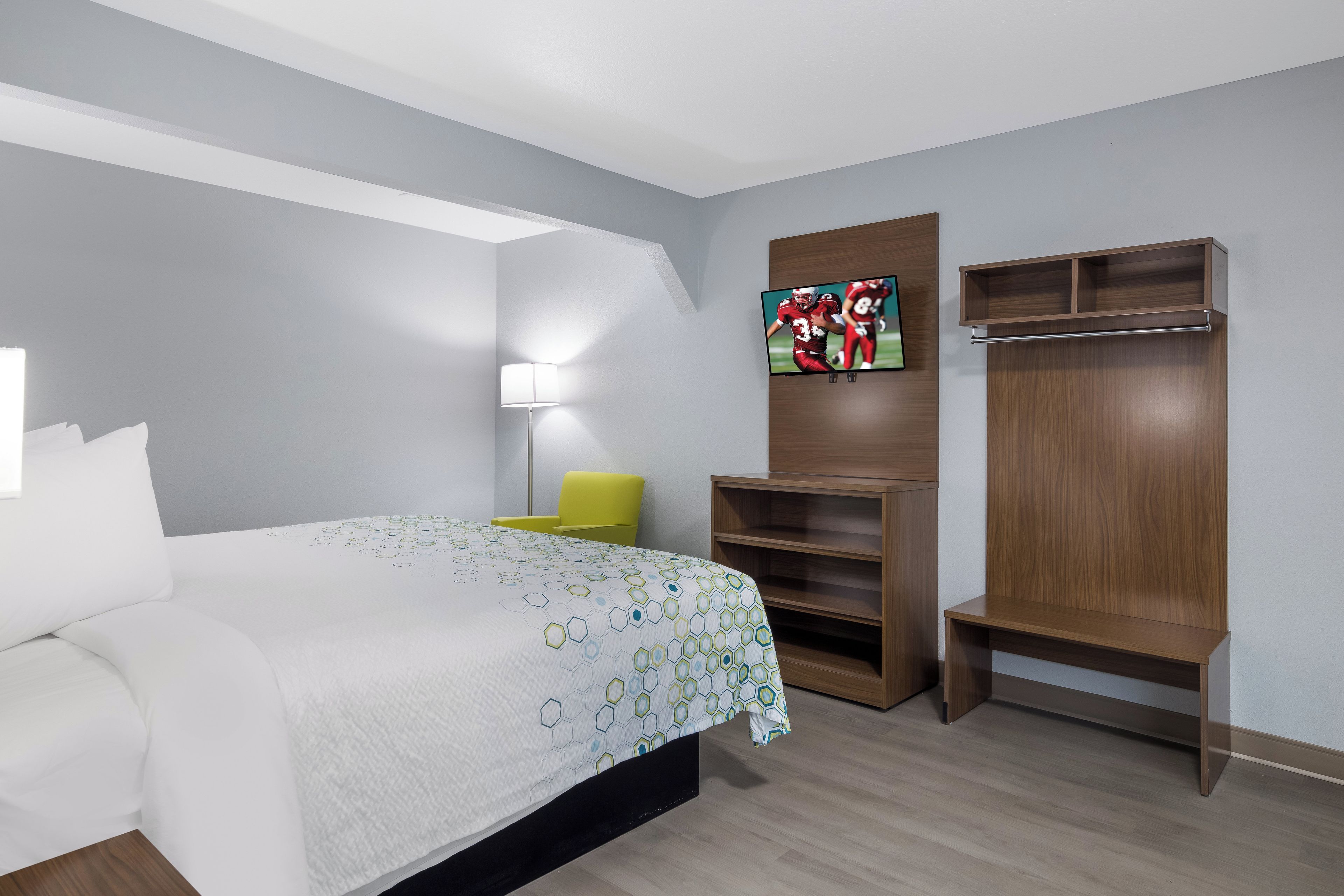 Quarto de hotel com uma cama, televisão fixada na parede, uma cadeira e um guarda-roupa de madeira.