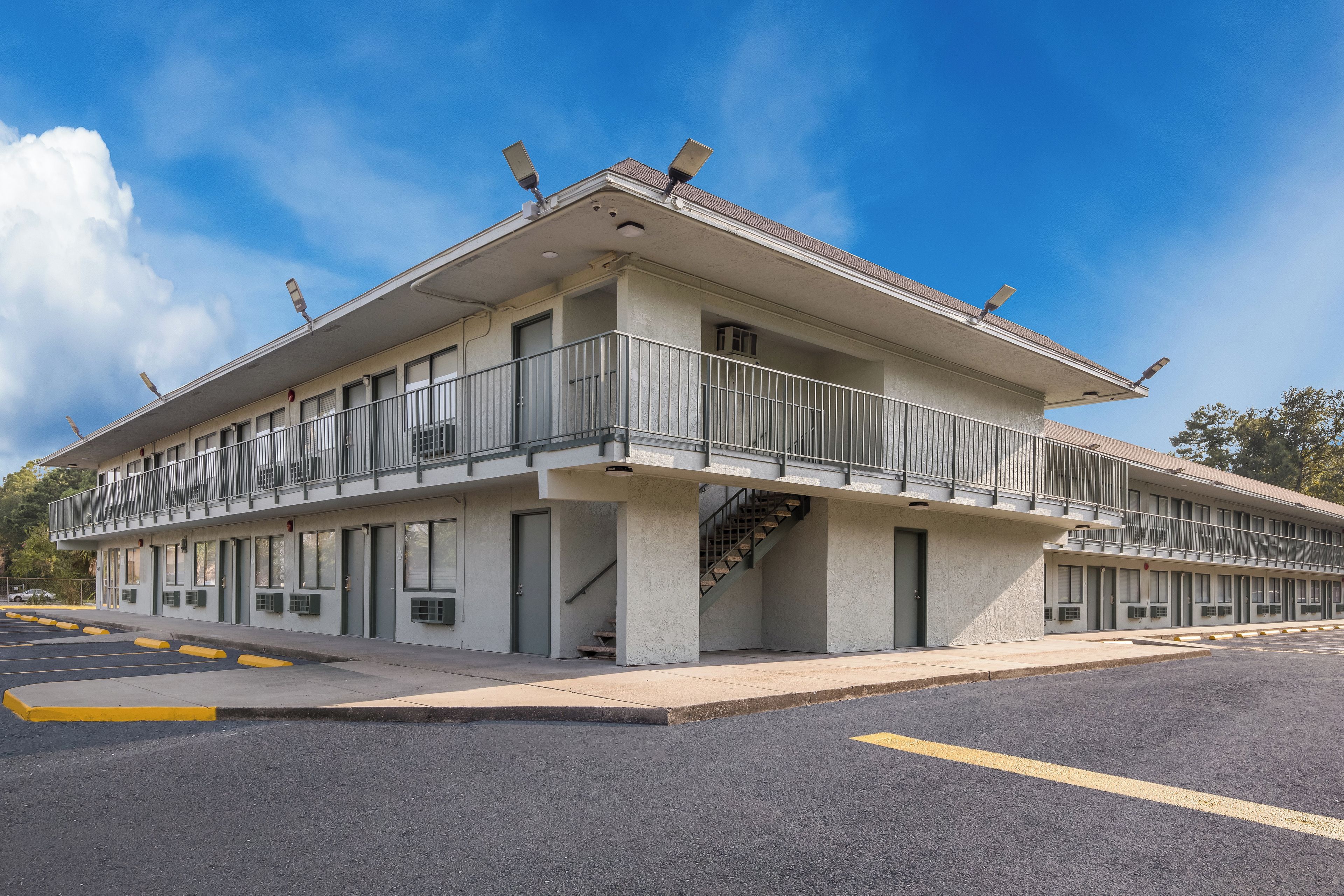 Um motel de dois andares com varanda no andar superior, escada que leva ao andar de cima e estacionamento na frente.