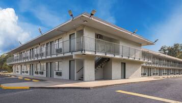 Um motel de dois andares com varanda no andar superior, escada que leva ao andar de cima e estacionamento na frente.