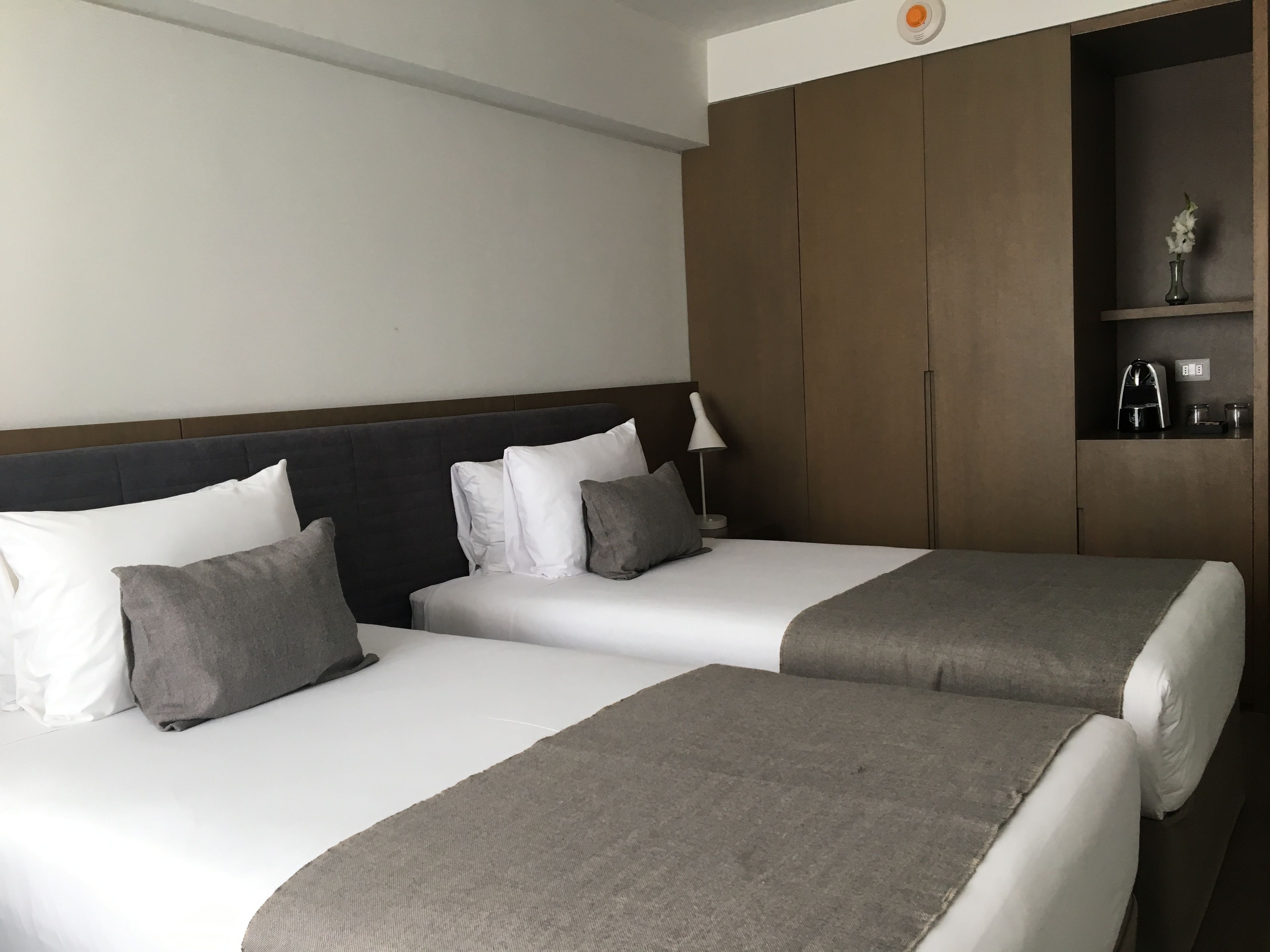 Quarto de hotel com duas camas, cada uma com lençóis brancos e cobertores cinzentos. Há uma mesa de cabeceira com abajur, um guarda-roupa de madeira e uma prateleira com um vaso e uma cafeteira.