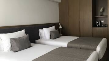 Quarto de hotel com duas camas, cada uma com lençóis brancos e cobertores cinzentos. Há uma mesa de cabeceira com abajur, um guarda-roupa de madeira e uma prateleira com um vaso e uma cafeteira.