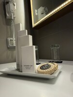 Casa de banho com lavatório branco, espelho com luzes LED, secador de cabelo e um porta-sabonetes com touca de banho, elástico de cabelo e barra de sabão.
