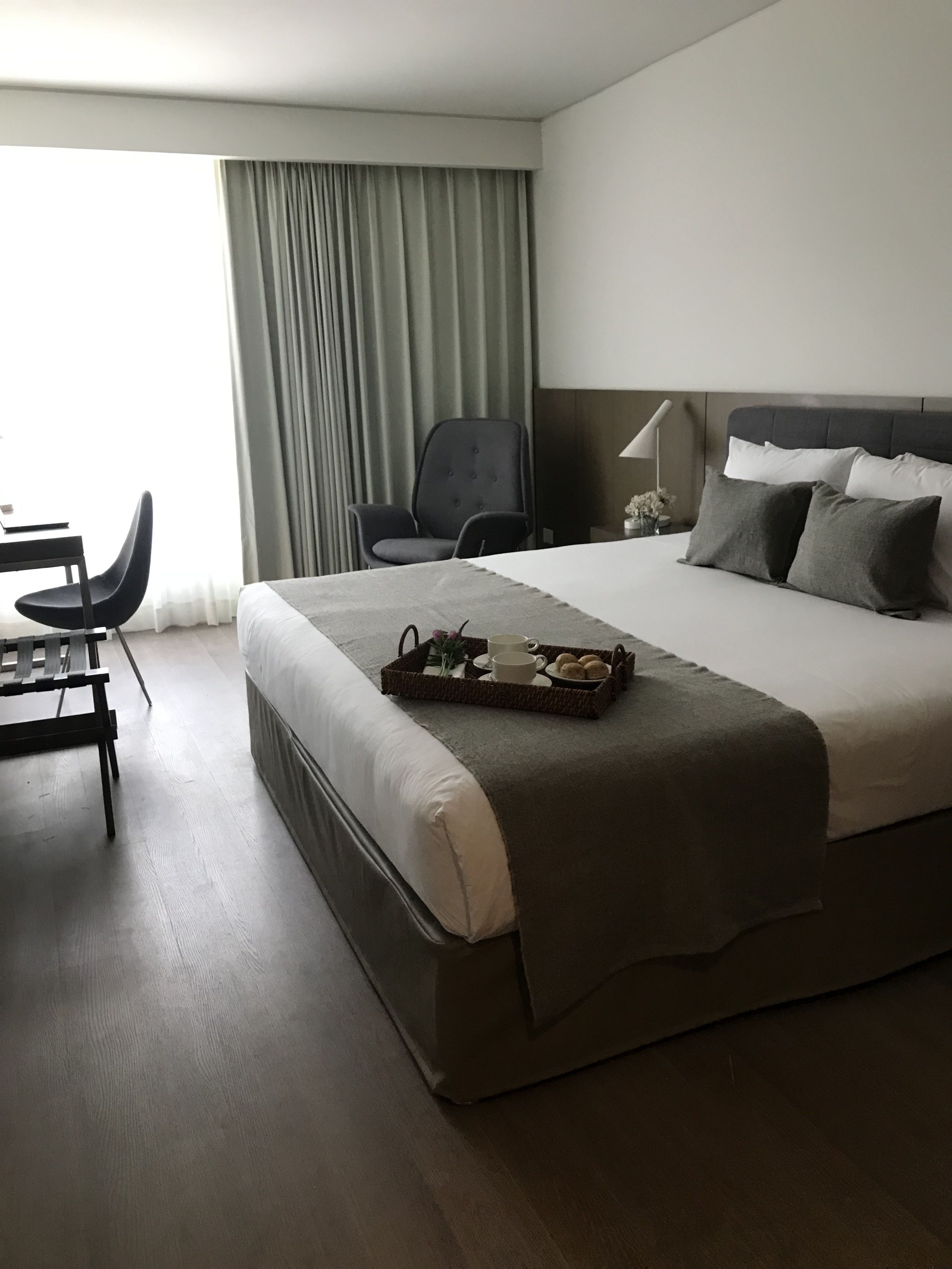 Quarto de hotel com uma cama grande, uma mesa com cadeira, uma luminária e uma janela com cortinas.