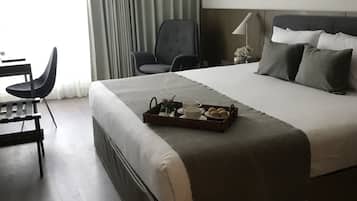 Quarto de hotel com uma cama grande, uma mesa com cadeira, uma luminária e uma janela com cortinas.