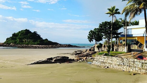 Uma praia com um muro de pedra, palmeiras e um edifício com fachada amarela.