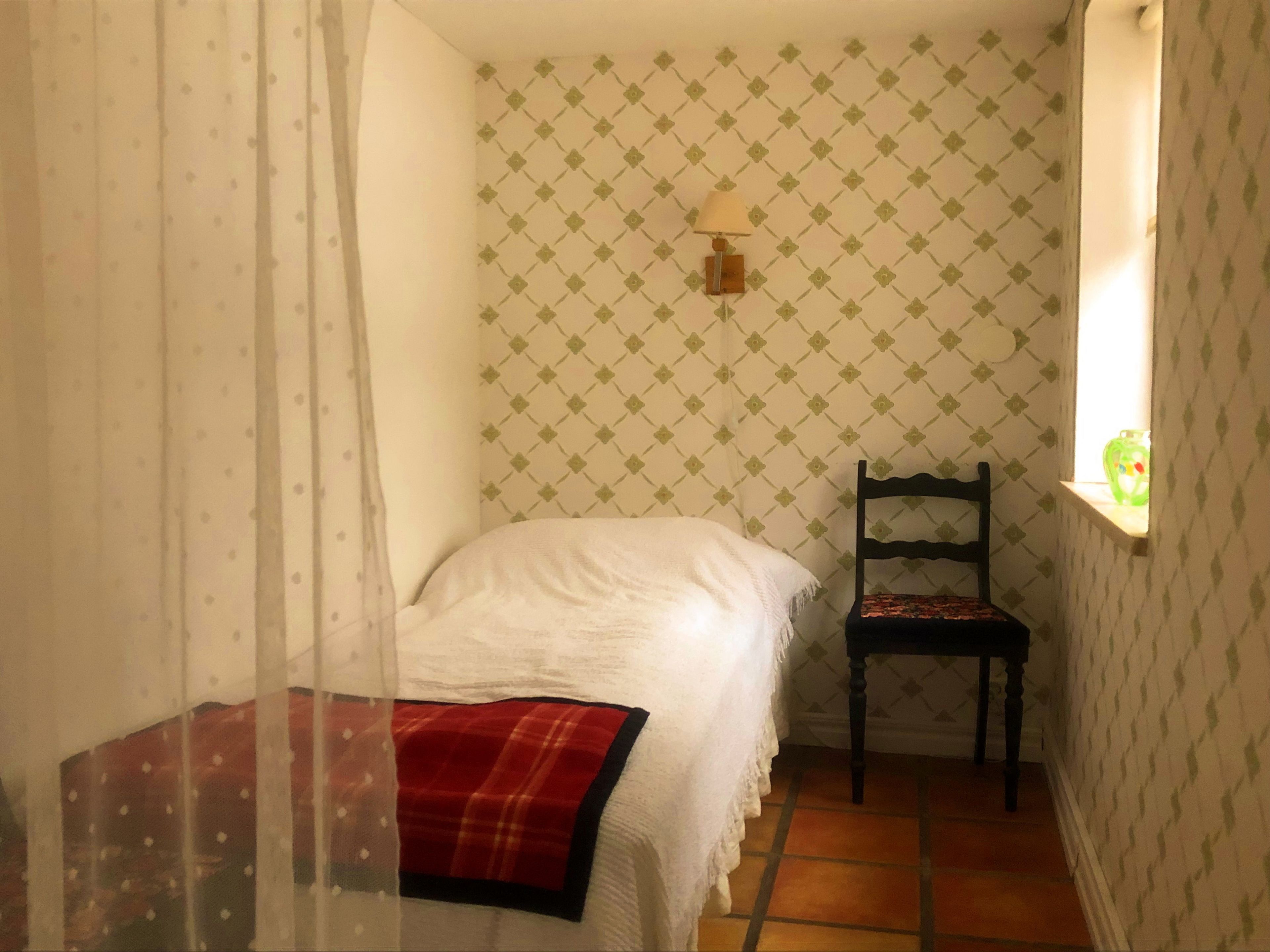 Quarto pequeno com cama, cadeira, luminária na parede e janela com vista.