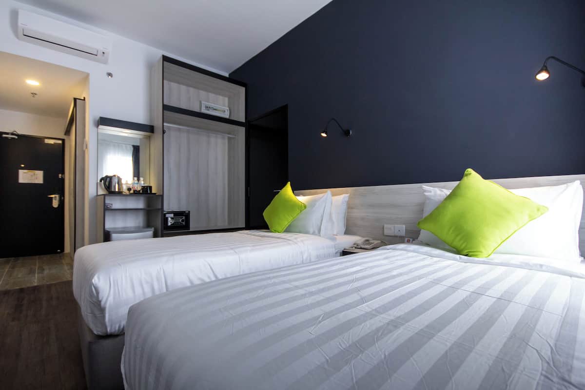 Quarto de hotel com duas camas, um guarda-roupa, um espelho e uma pequena escrivaninha com abajur.