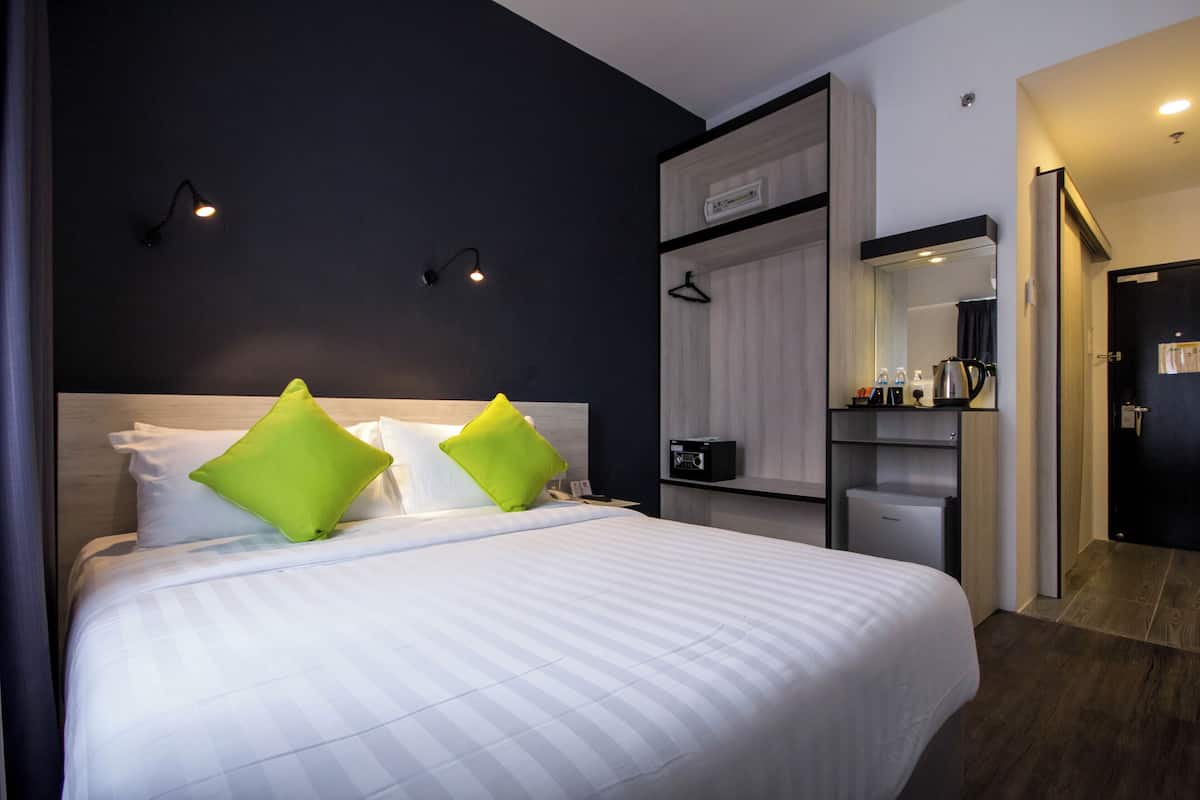 Quarto de hotel com uma cama grande, duas almofadas verdes, uma mesa de cabeceira com abajur, um guarda-roupa e uma pequena cozinha.