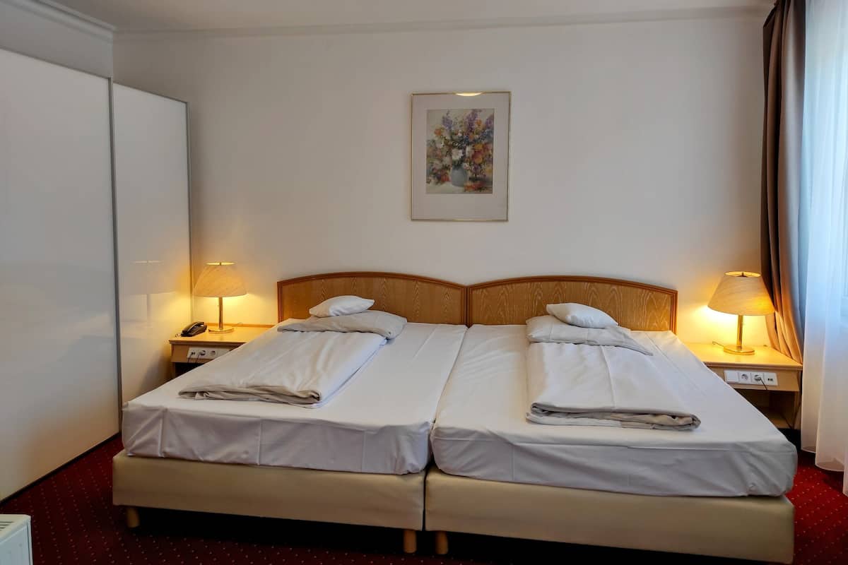 Uma cama de casal com roupa de cama branca, duas mesas de cabeceira com abajures, uma obra de arte floral emoldurada na parede e um chão coberto de carpete vermelho.