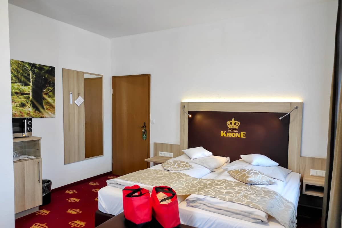 Quarto de hotel com uma cama, uma cabeceira com o logotipo de uma coroa, um tapete vermelho e um par de malas vermelhas sobre uma mesa.