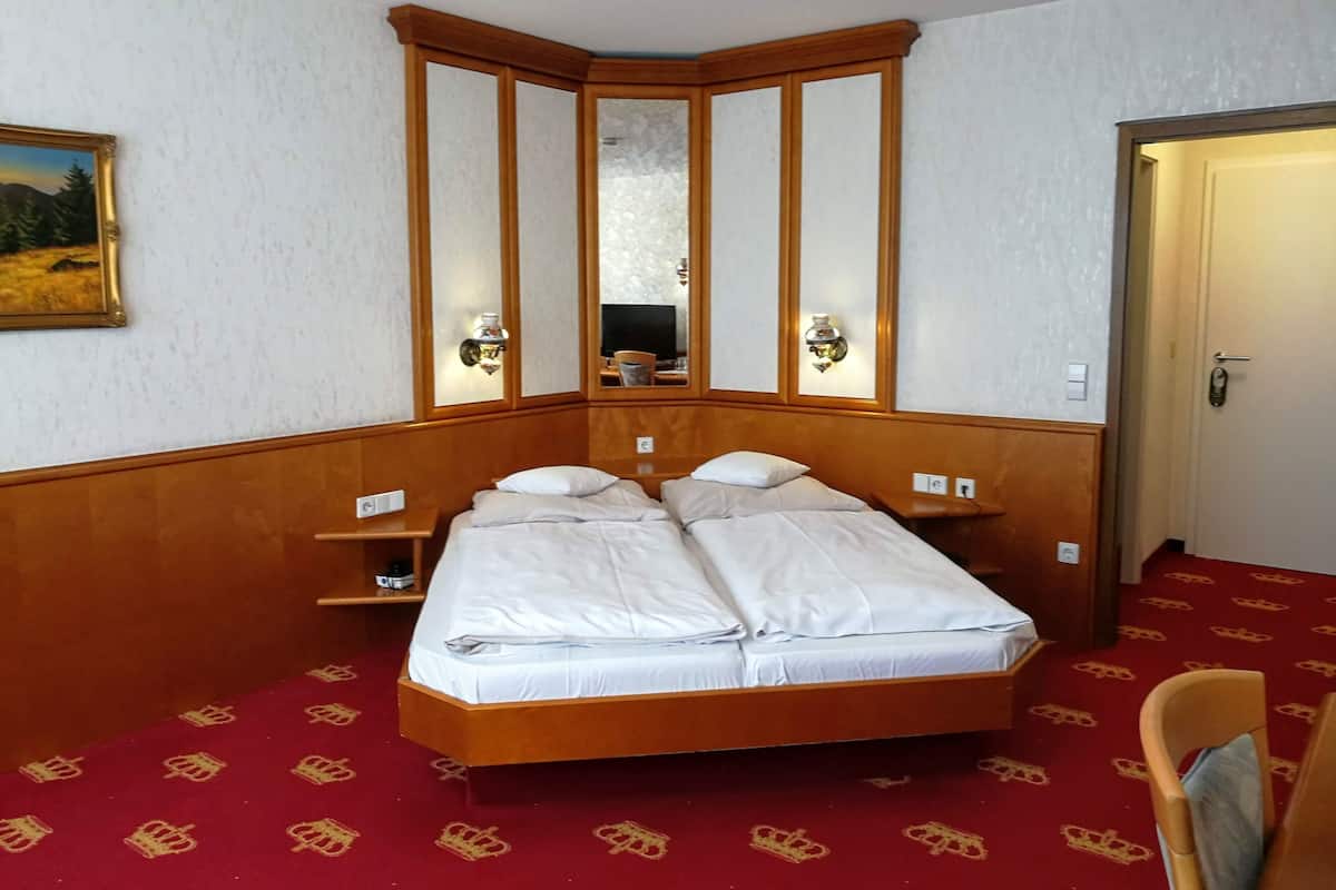 Quarto de hotel com cama de canto, um quadro na parede e um tapete vermelho com padrão de coroa.
