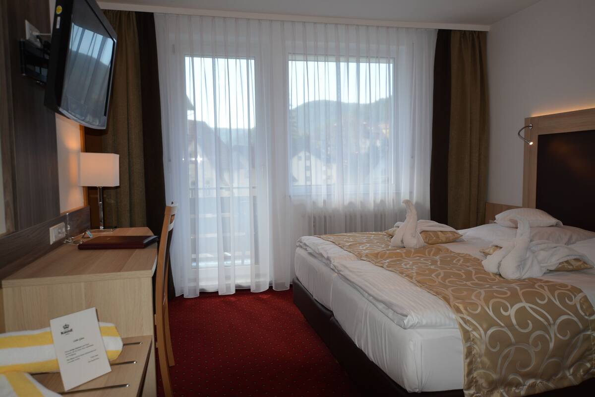Quarto de hotel com cama, escrivaninha com luminária, televisão e vista para o exterior através de cortinas translúcidas.