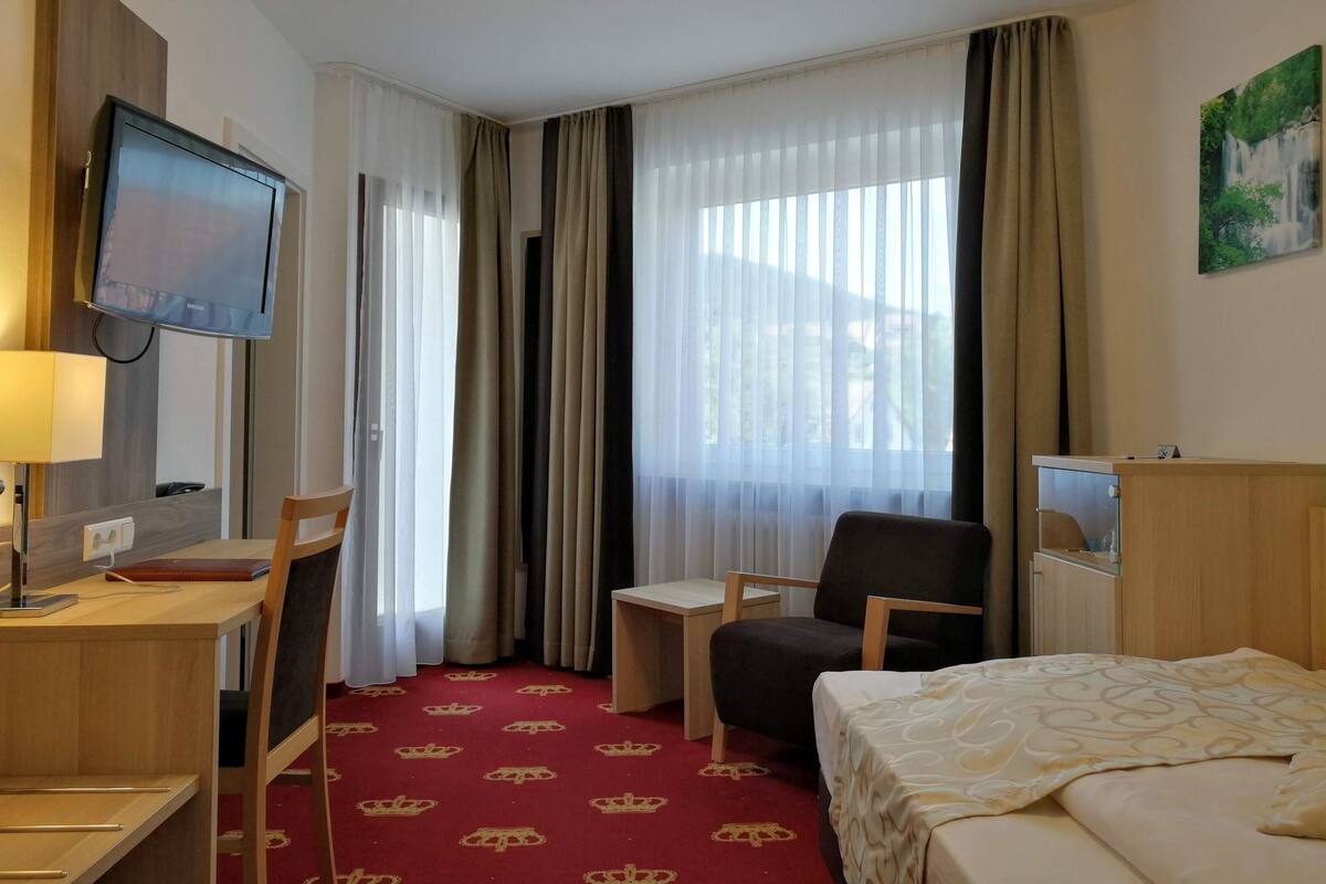 Quarto de hotel com uma cama, uma escrivaninha com cadeira, uma televisão e uma janela com cortinas.