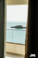 Vista da janela com cortina, sobre um mar calmo e um afloramento rochoso.