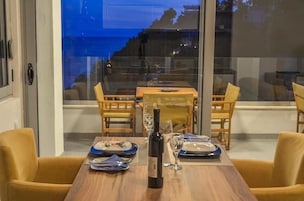 Uma área de refeições com uma mesa posta para dois, uma garrafa de vinho e vista para o mar através de grandes janelas.
