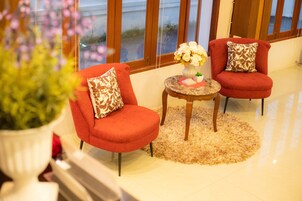Quarto com duas poltronas vermelhas, uma mesa redonda pequena com um vaso de flores e uma janela grande com persianas.