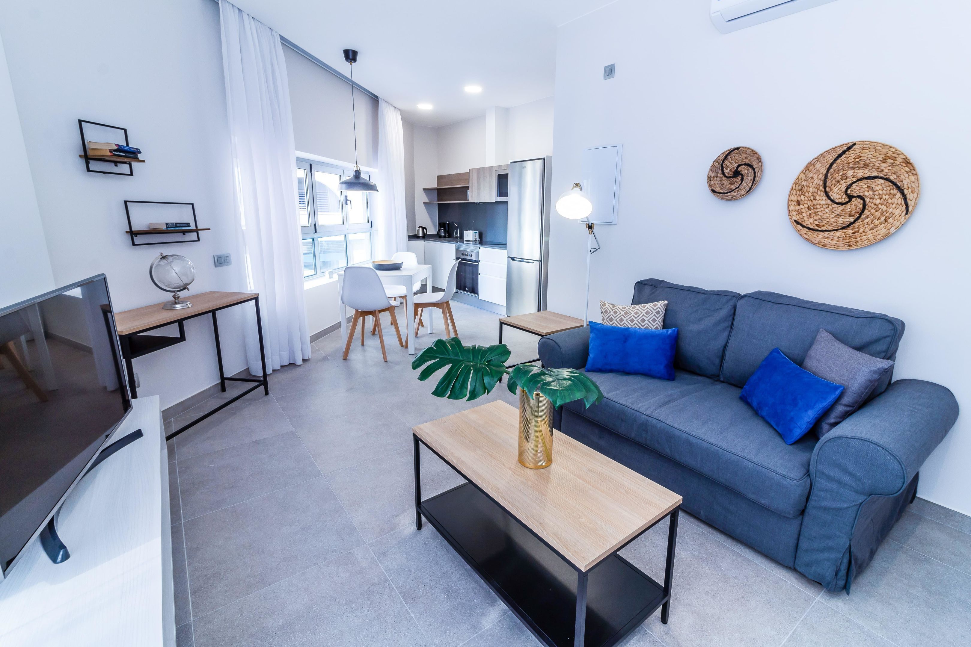 Uma sala de estar moderna com um sofá azul, uma mesa de centro e uma televisão. Uma cozinha com área de refeições está visível ao fundo.